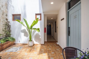 Um corredor estreito com piso ladrilhado, um vaso de planta e uma porta com placa de número.