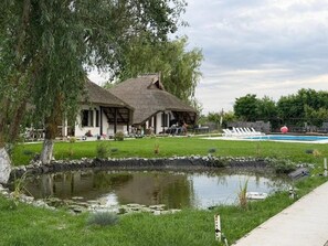 Uma casa com telhado de palha, um lago e uma piscina ao fundo.