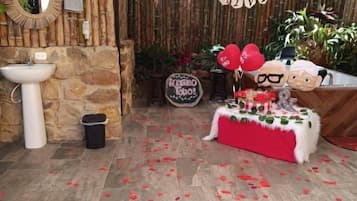 Ambiente rústico ao ar livre com parede de pedra, um banco de madeira e uma mesa com toalha vermelha.