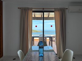 Quarto com mesa e cadeiras, uma garrafa de água e vista para o mar através de uma porta de vidro.