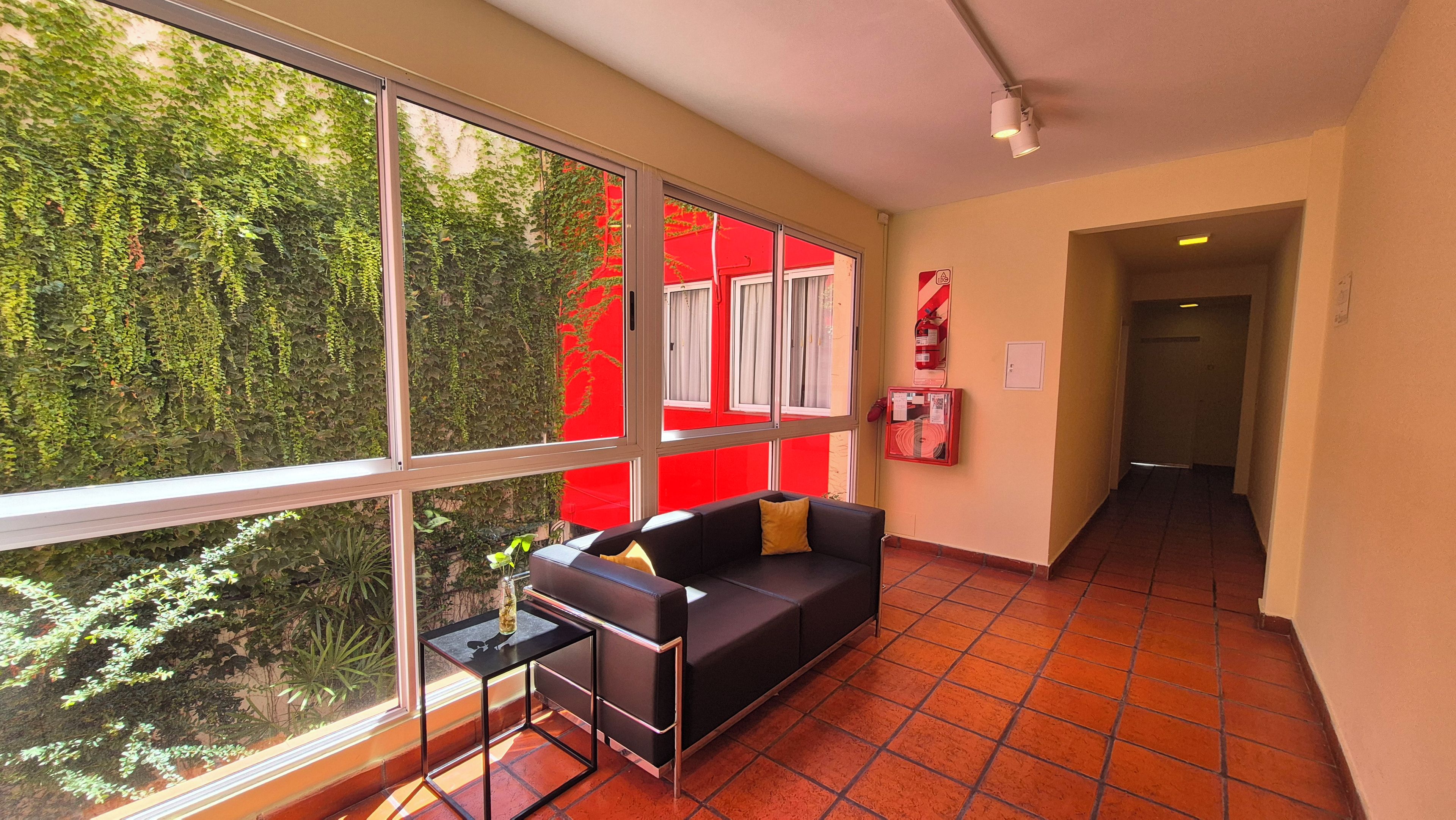 Quarto com sofá preto, mesa pequena e grande janela com vista para área verde.