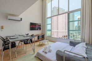 Quarto de hotel moderno com uma janela grande, cama, escrivaninha com cadeiras, TV de tela plana e ar condicionado.