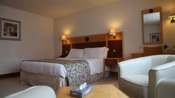 Quarto de hotel com uma cama, mesas de cabeceira, uma cadeira, uma mesinha com um estojo de lenços de papel e um espelho na parede.