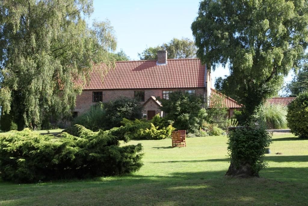 Uma casa de tijolos com telhado de telhas vermelhas, rodeada por um gramado bem cuidado e diversas árvores.