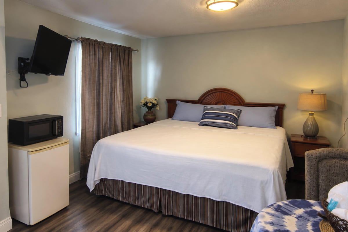 Quarto de hotel com cama, televisão na parede, secretária pequena com abajur e uma cadeira.