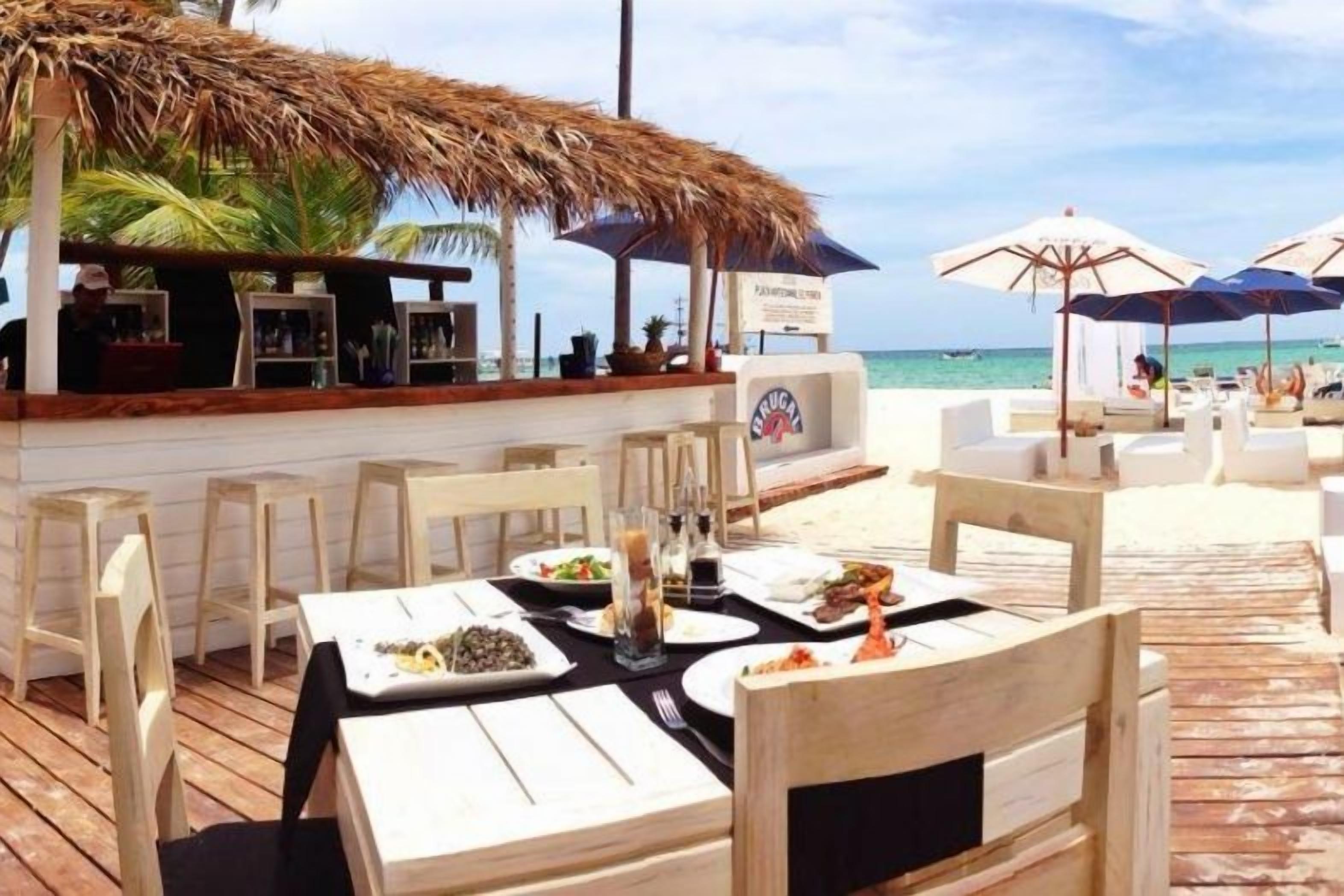 Restaurante à beira-mar com mesas e cadeiras de madeira, área de bar e guarda-sóis para proteção do sol.