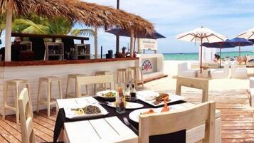 Restaurante à beira-mar com mesas e cadeiras de madeira, área de bar e guarda-sóis para proteção do sol.