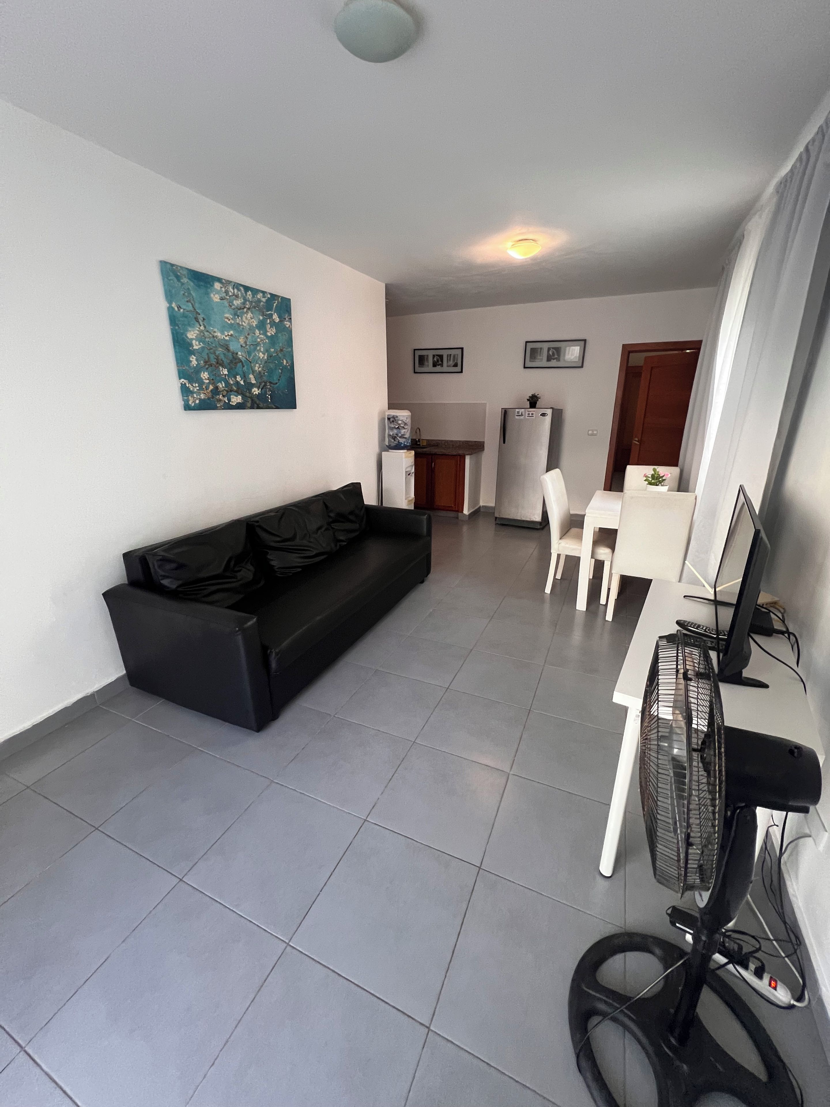 Uma sala de estar moderna com um sofá preto, uma área de jantar com mesa branca e uma cozinha com geladeira e armários.