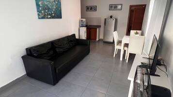 Uma sala de estar moderna com um sofá preto, uma área de jantar com mesa branca e uma cozinha com geladeira e armários.