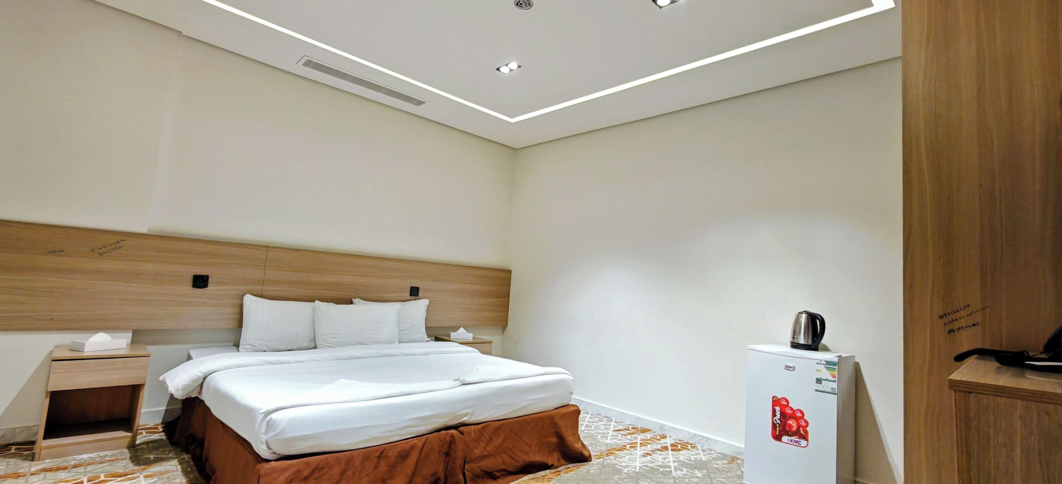 Quarto de hotel com cama, mesa de cabeceira, escrivaninha com chaleira e geladeira.