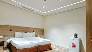 Quarto de hotel com cama, mesa de cabeceira, escrivaninha com chaleira e geladeira.