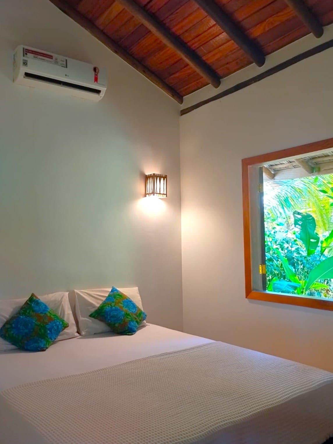 Quarto com cama, almofadas, ar condicionado, luminária na parede e janela vista para vegetação.