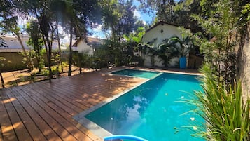 Piscina com deck de madeira, rodeada por vegetação exuberante e um edifício com telhado telhado.