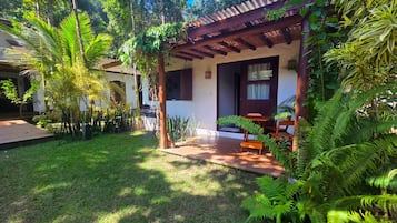 Uma área de estar coberta ao ar livre com mesa e cadeiras, cercada por vegetação exuberante e um gramado bem cuidado.
