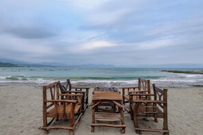 Uma praia com cadeiras e mesa de madeira, com vista para um mar calmo e barcos.