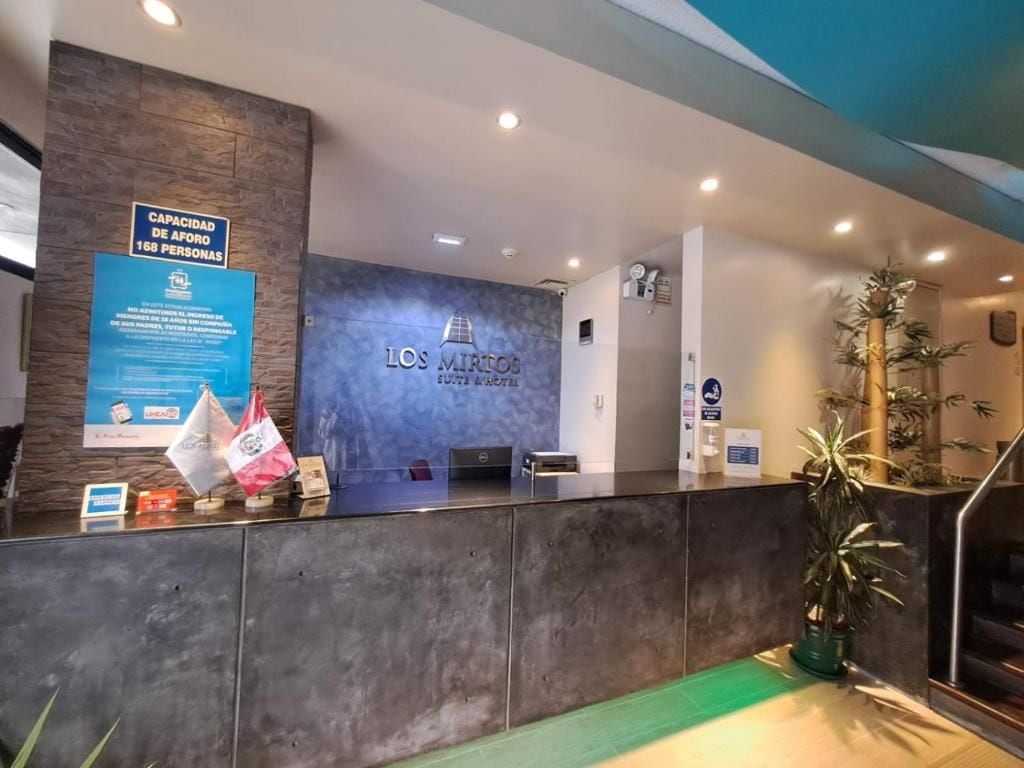 Uma área de recepção com placa de capacidade, parede azul com o nome do hotel, balcão de atendimento e um vaso de planta.