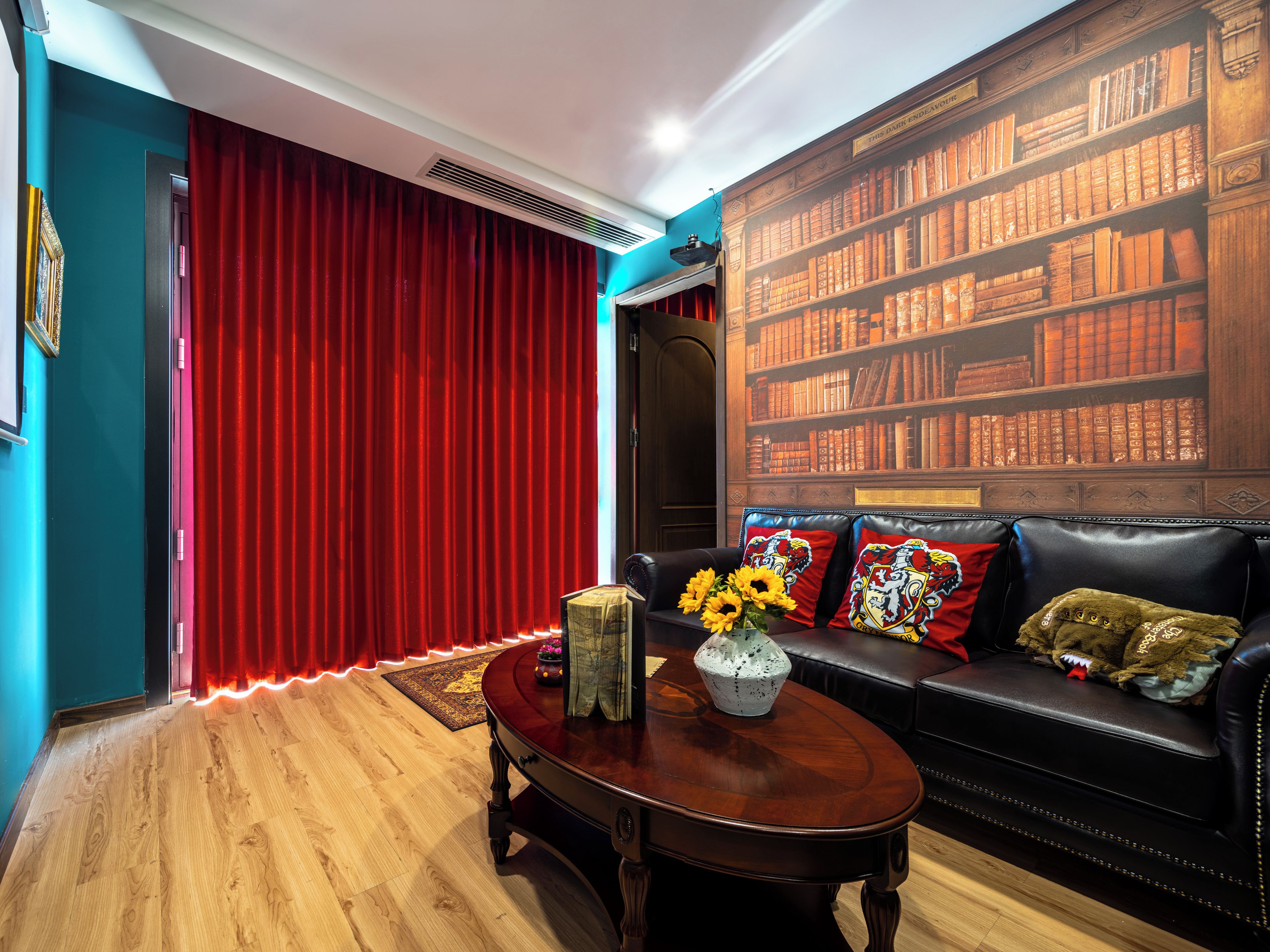 Um quarto com piso de madeira, um sofá de couro preto com almofadas decorativas, uma mesa de centro de madeira com um vaso de girassóis e uma estante cheia de livros.