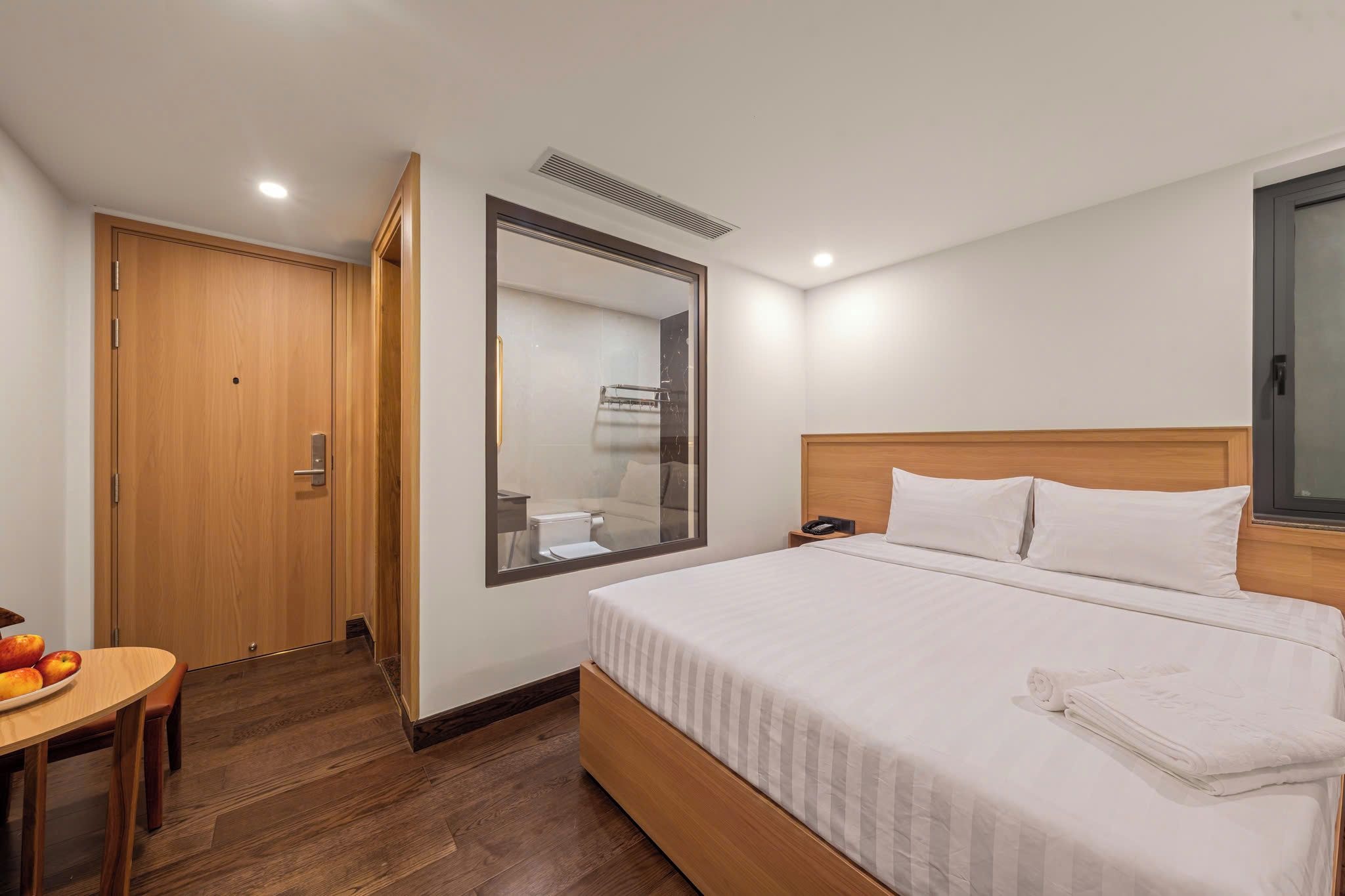 Quarto de hotel com cama, porta de madeira, espelho de banheiro e uma pequena mesa com frutas.