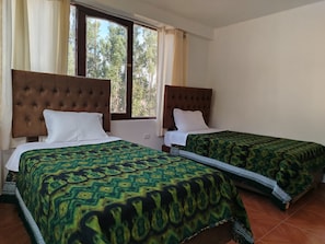 Quarto de hotel com duas camas, cada uma com cabeceira estofada, colcha estampada e janela com cortinas.