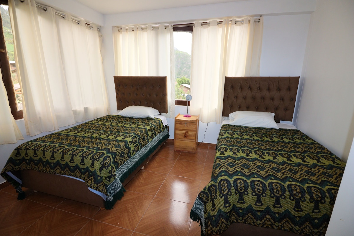 Quarto de hotel com duas camas, cada uma com colcha estampada, uma mesinha de cabeceira de madeira e vista para o verde lá fora.