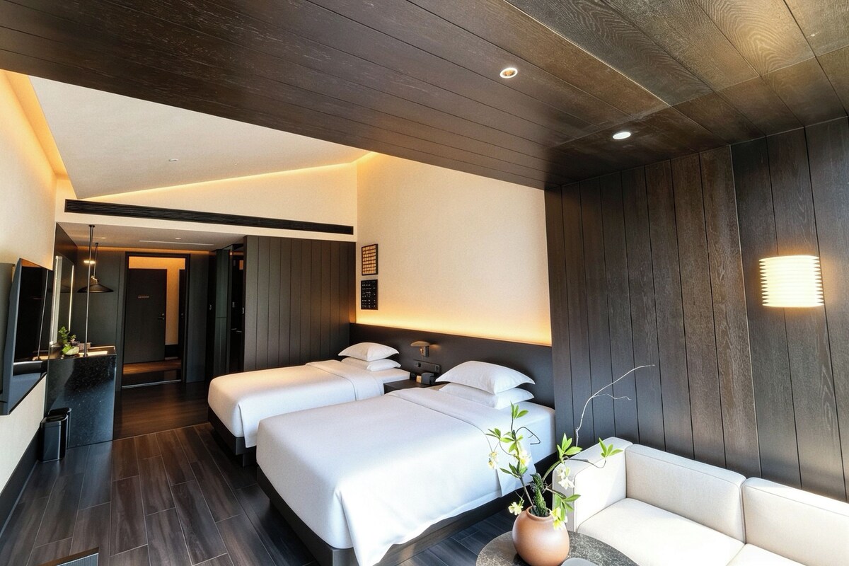 Quarto de hotel moderno com duas camas, um sofá, um vaso de plantas e um painel de controle fixado na parede.