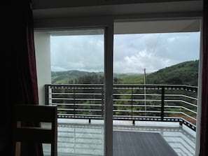 Quarto com uma grande porta de vidro deslizante que dá para uma varanda com guarda-corrimão de metal, oferecendo vista para colinas verdes e um céu nublado.