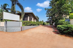 Uma entrada pavimentada conduz a um edifício moderno com telhado inclinado, cercado por vegetação exuberante e palmeiras.