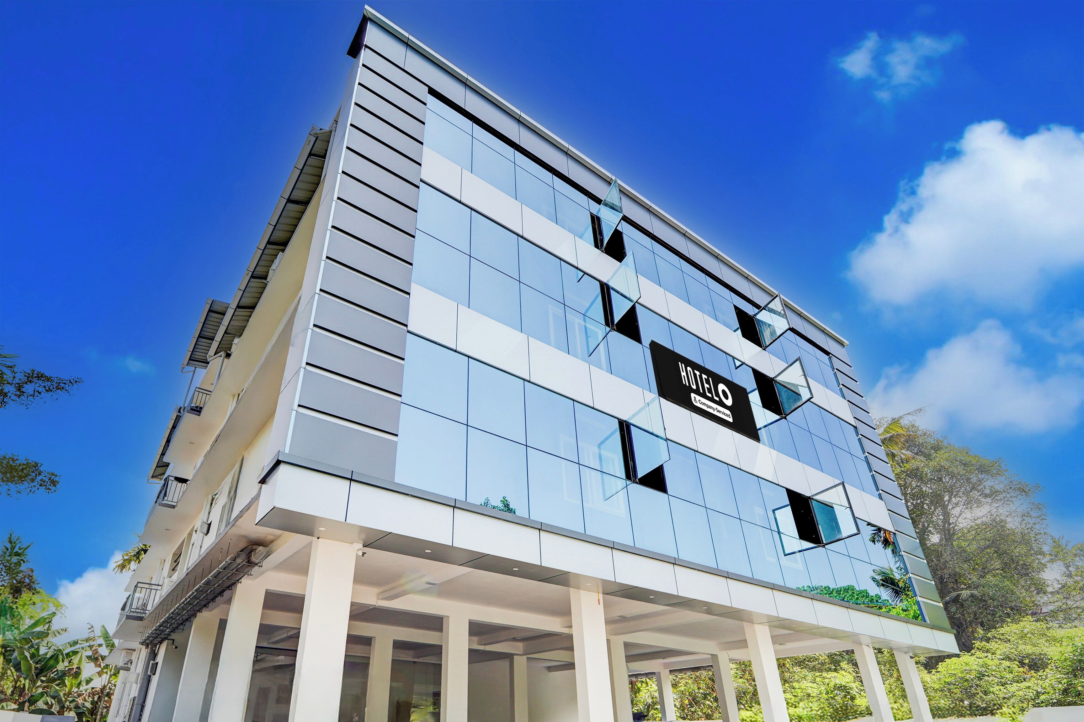Um edifício moderno de hotel com fachada de vidro, placa indicando ser um hotel de propriedade da empresa, e área de entrada coberta.
