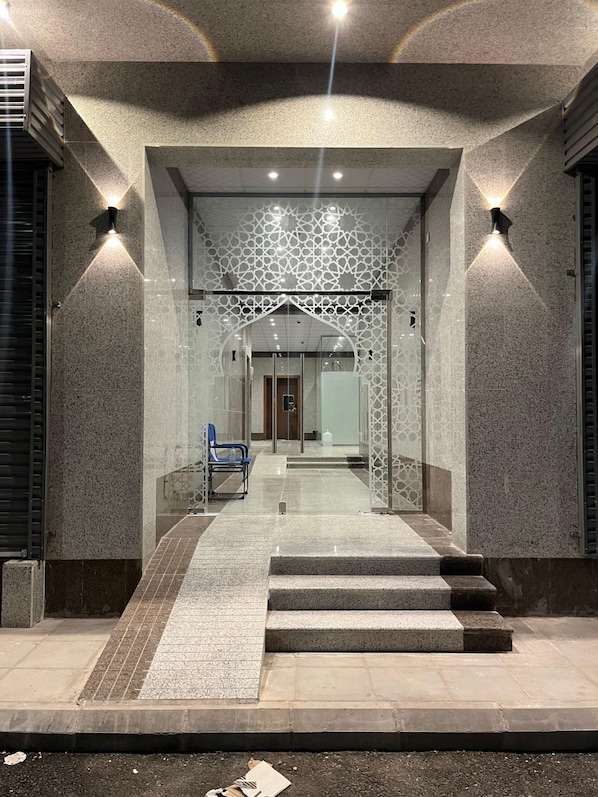 Entrada de um edifício moderno com uma parede de padrão geométrico, uma cadeira azul e um conjunto de escadas que levam à entrada.