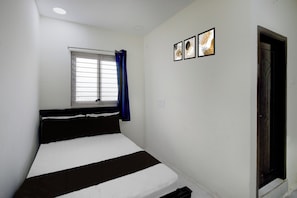 Quarto com uma cama de solteiro, janela com persianas, uma porta e três quadros na parede.