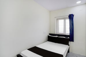 Uma cama de solteiro com colcha preta e branca em quarto com paredes brancas e janela com cortinas azuis.