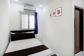 Uma cama de solteiro com colcha listrada em preto e branco, uma janela com persianas, uma cortina azul e uma porta com puxador decorativo.