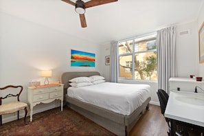 Quarto com cama, escrivaninha, cadeira, mesa de cabeceira, ventilador de teto, janela com cortinas e um quadro na parede.
