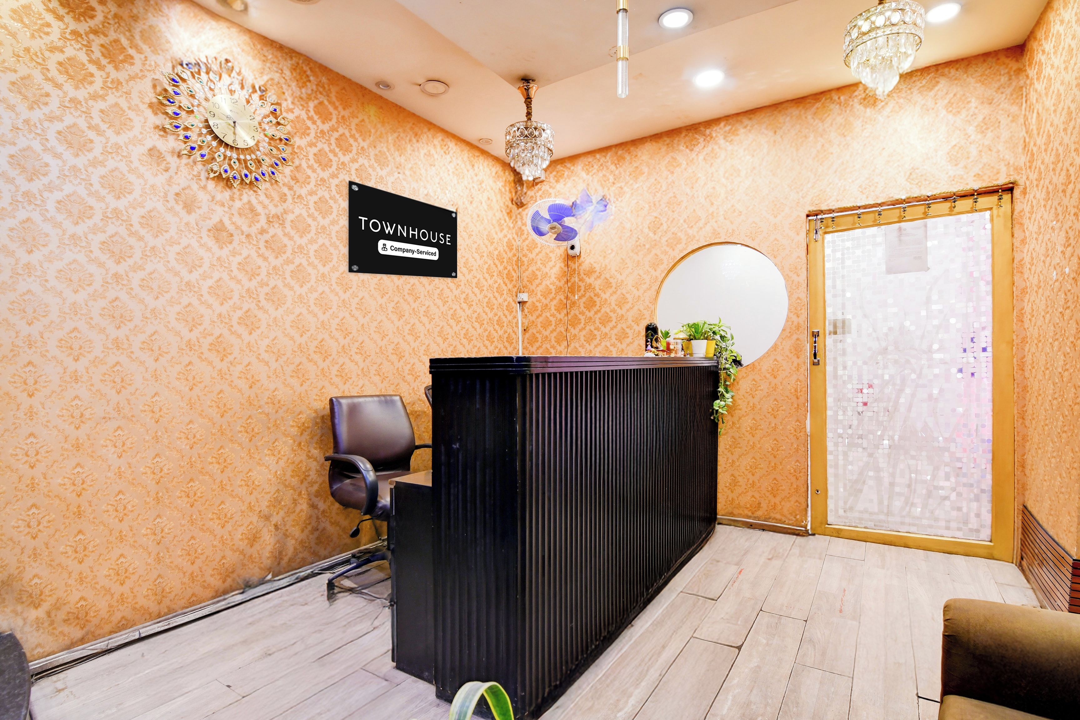 Uma área de recepção com um balcão preto, uma placa com a inscrição 'TOWNHOUSE Company Serviced', um ventilador, um espelho e uma porta com padrão de mosaico.