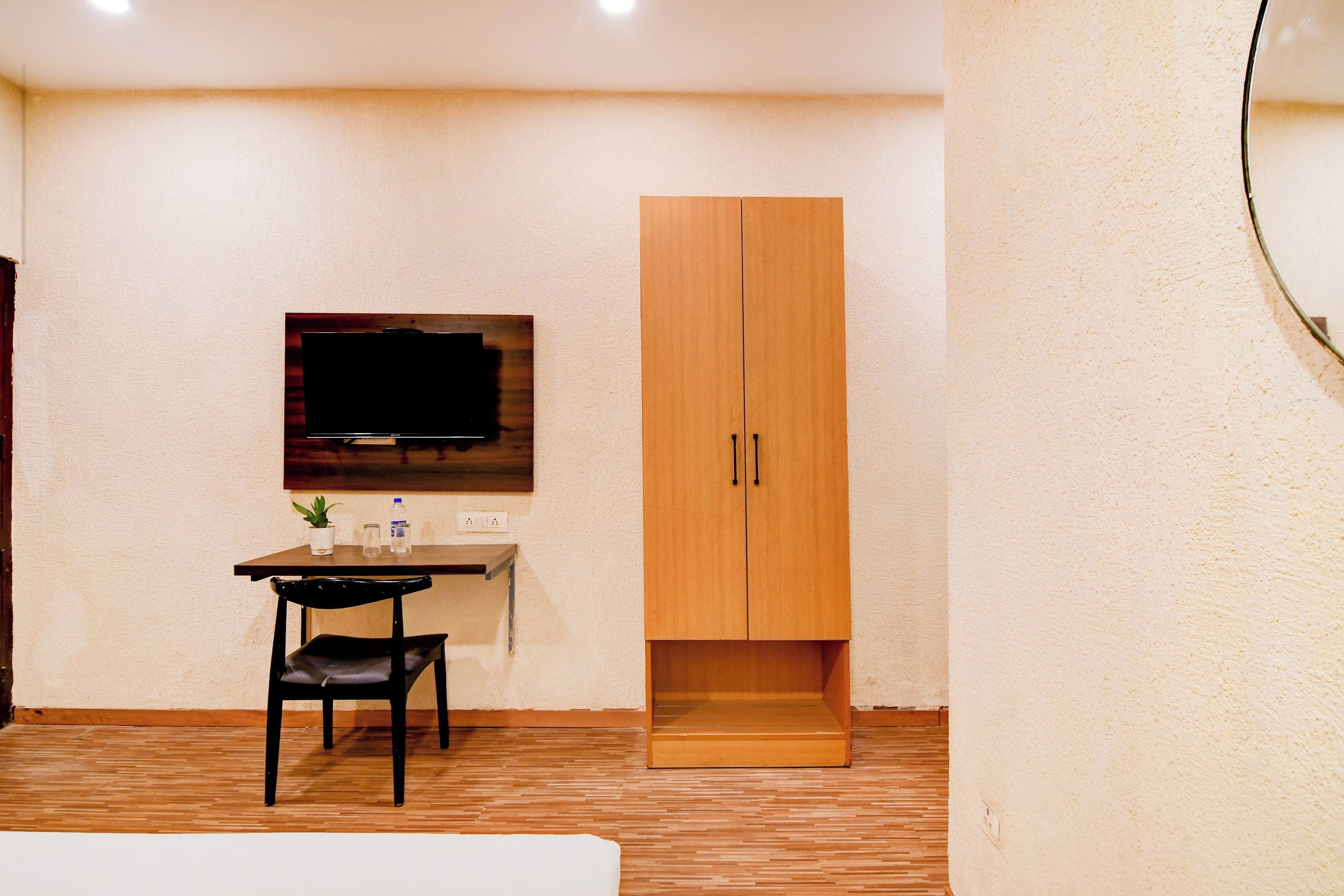 Quarto com guarda-roupa de madeira, televisão na parede, mesa pequena com cadeira e planta em vaso.