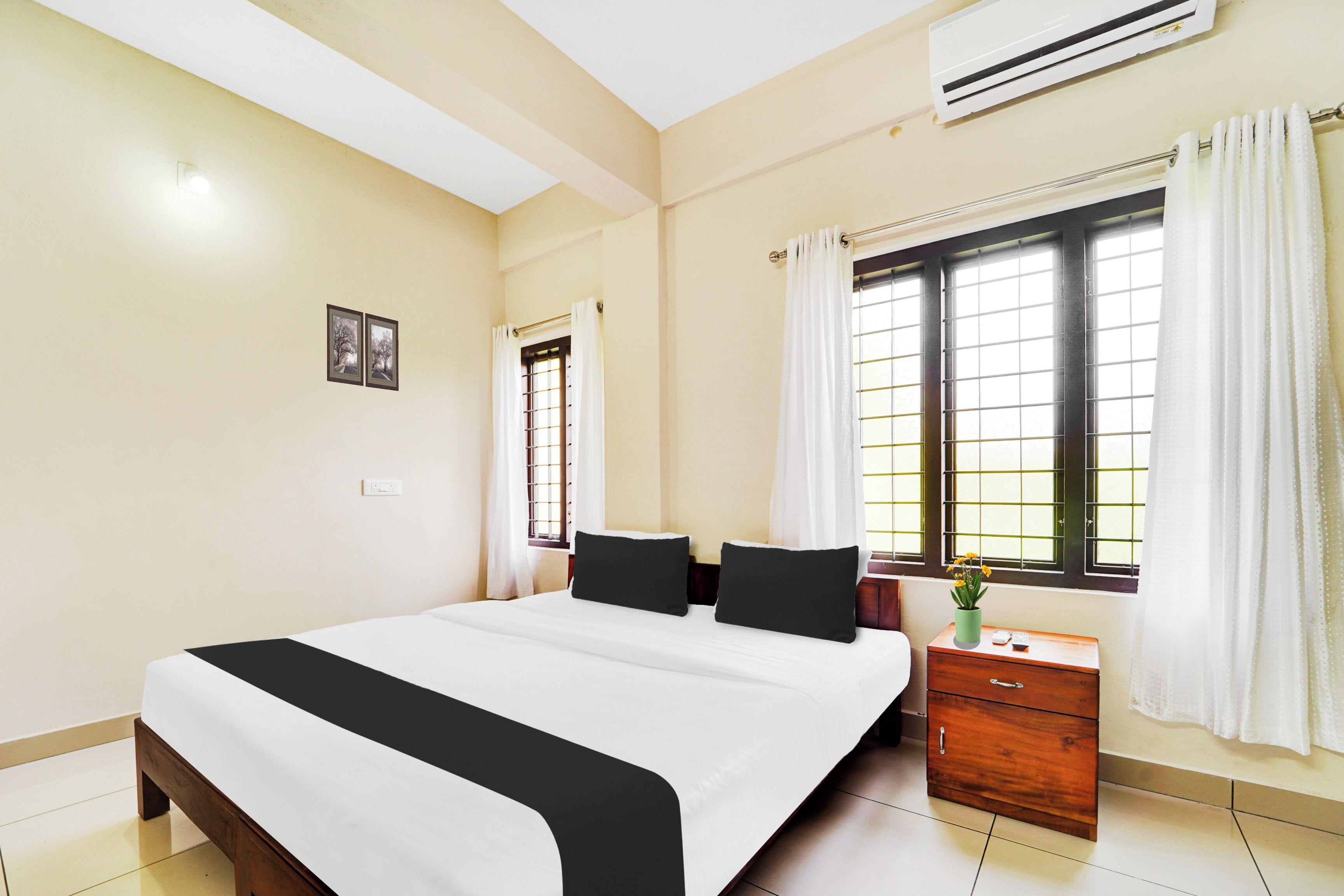 Quarto de hotel com uma cama grande, uma mesa de cabeceira com um vaso de flores e uma janela com cortinas.