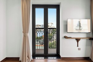 Entrada de um quarto de hotel com porta de vidro emoldurada a preto, cortinas bege, soalho de madeira e um quadro com a imagem da House of Hanoi na parede.