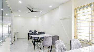 Sala de conferências com mesa branca, cadeiras cinzas, ventilador de teto e janela com padrão quadriculado.