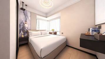 Quarto de hotel moderno com uma cama grande, uma escrivaninha com cadeira, dispensador de água e televisão fixada na parede.