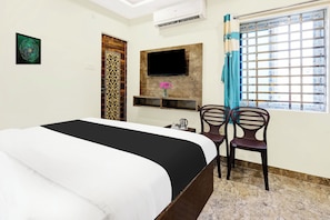 Quarto de hotel com cama, televisão de tela plana na parede, mesa pequena com um vaso de flores, duas cadeiras e janela com cortinas.