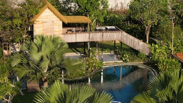Uma área de piscina com estrutura de telhado de palha, rodeada por vegetação exuberante e uma ponte de madeira.