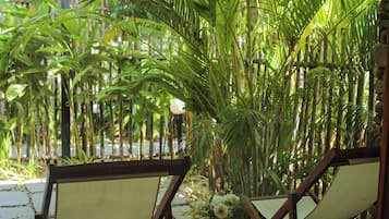 Vista pela janela, com uma mesa e cadeiras, em meio a plantas verdes exuberantes.
