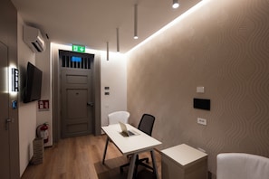 Quarto de hotel moderno com piso de madeira, uma escrivaninha com um laptop, uma cadeira, uma televisão e um extintor de incêndio.