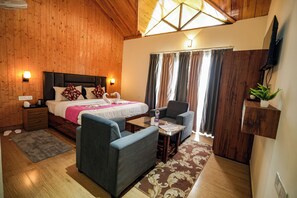 Quarto de hotel com teto de madeira, cama com almofadas, mesa pequena com uma garrafa, cadeira e televisão fixada na parede.
