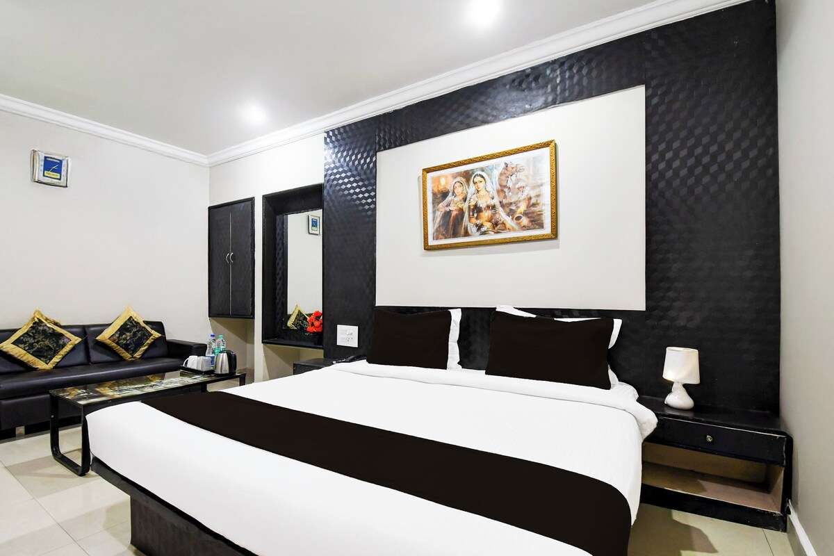 Quarto de hotel com uma cama grande, esquema de cores preto e branco, um quadro na parede e uma pequena mesa com chaleira e garrafas de água.
