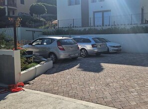 Uma área de estacionamento com três carros alinhados, um caminho pavimentado e um edifício ao fundo.