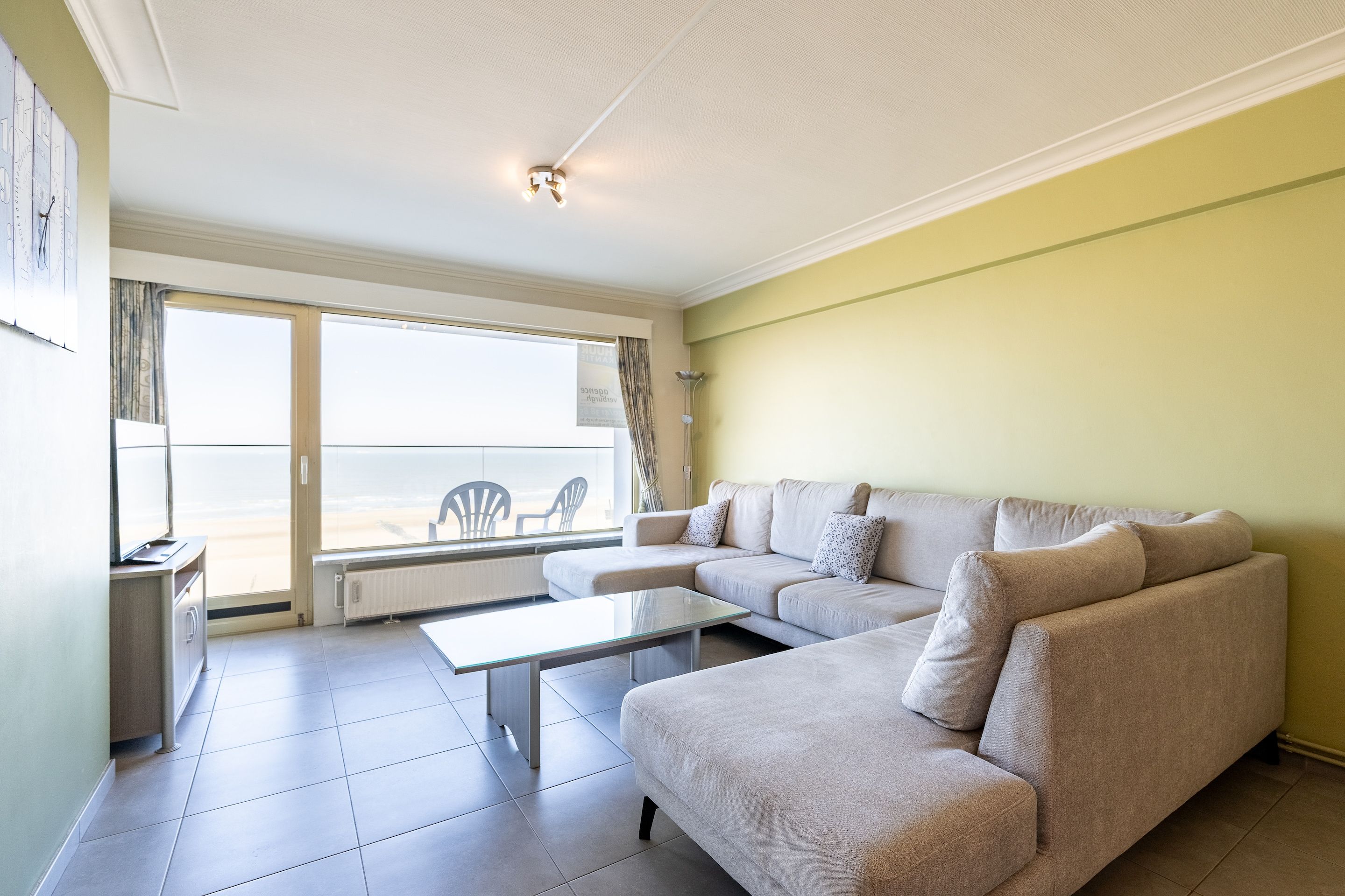 Sala de estar moderna com sofá, mesa de centro e vista para a praia através de grandes janelas.