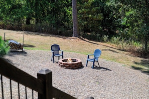 Um pátio de cascalho com duas cadeiras Adirondack, uma lareira e uma pilha de lenha.