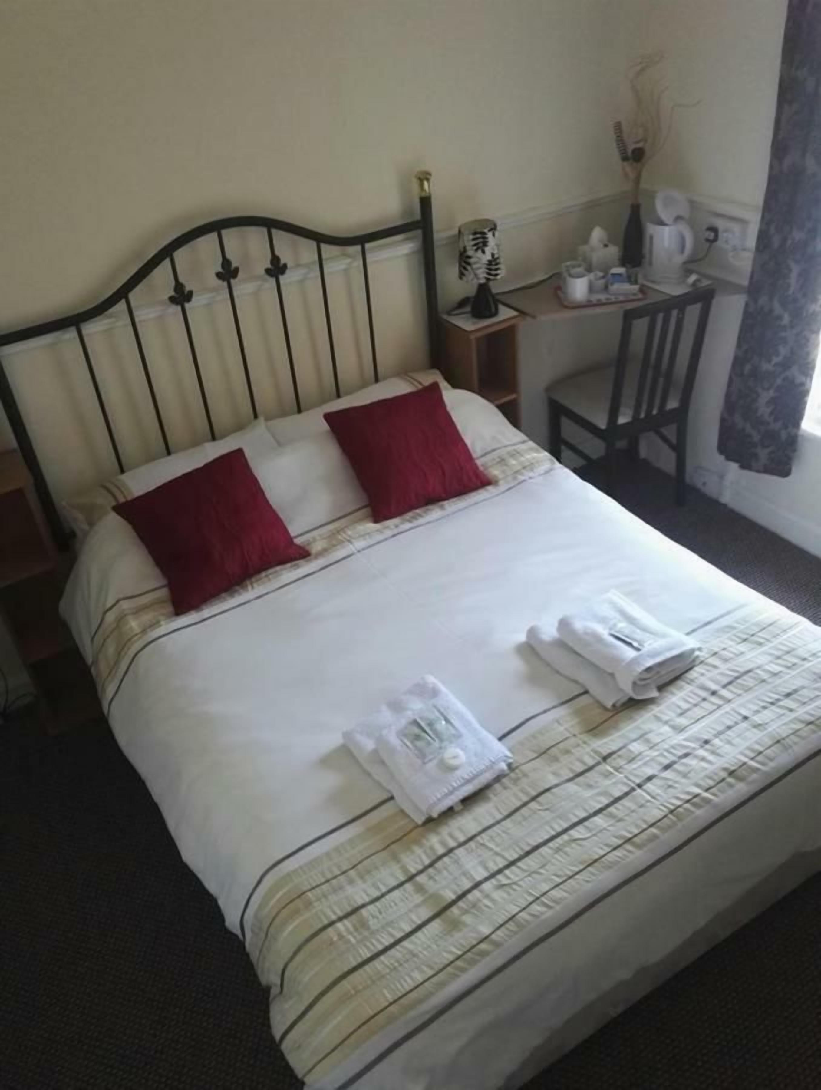 Cama bem arrumada com colcha listrada, dois travesseiros vermelhos e um conjunto de toalhas dobradas sobre a cama.