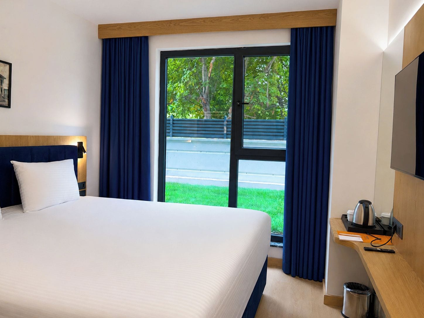Quarto de hotel com cama, escrivaninha com chaleira e vista para o verde lá fora.